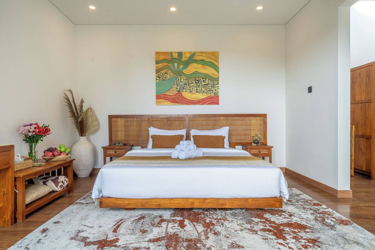 Uma cama grande com estrutura de madeira, mesa de cabeceira, um vaso com flores e um quadro na parede.