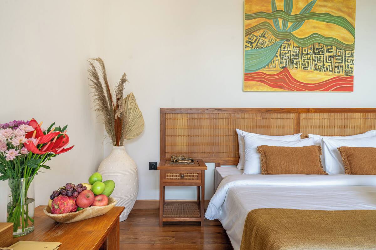 Quarto de hotel com cama, mesa de cabeceira, um vaso com flores, uma fruteira e uma pintura na parede.