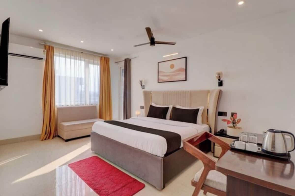 Quarto de hotel com cama, escrivaninha, cadeira, televisão, ventoinha no teto e janela com cortinas.
