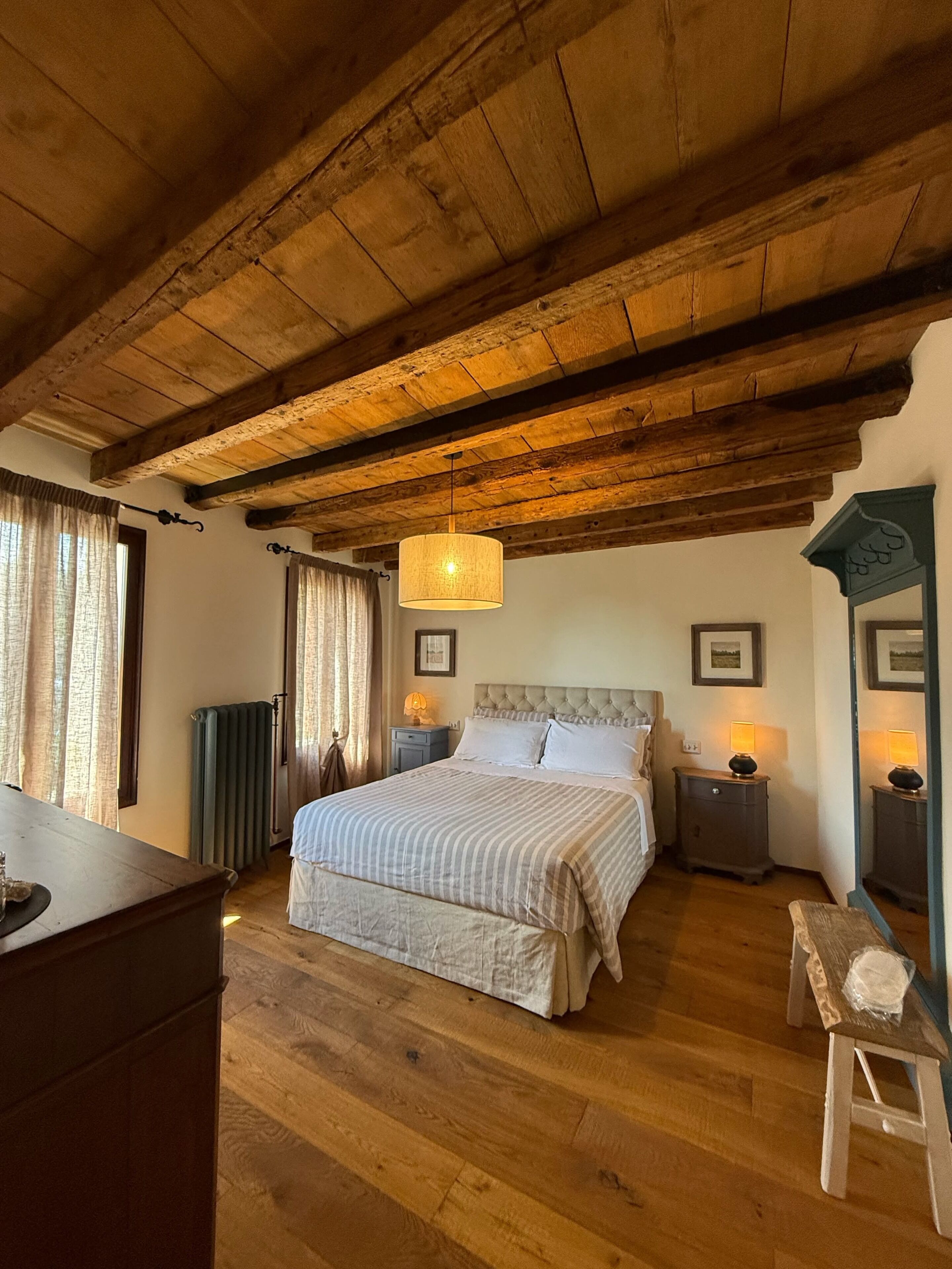 Quarto com teto de madeira, cama com colcha listrada, duas mesas de cabeceira com luminárias, cômoda e espelho.