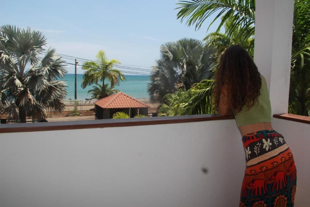 Uma varanda com vista para o mar, palmeiras e um pequeno edifício.