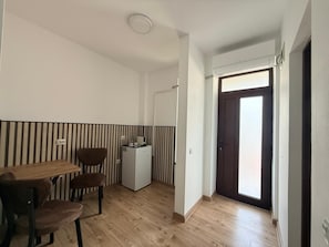 Um quarto pequeno com uma mesa de madeira, duas cadeiras, um refrigerador branco, uma parede listrada e uma porta que dá para o exterior.