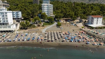 Vista aérea de uma praia com vários guarda-sóis e espreguiçadeiras, cercada por edifícios e árvores.