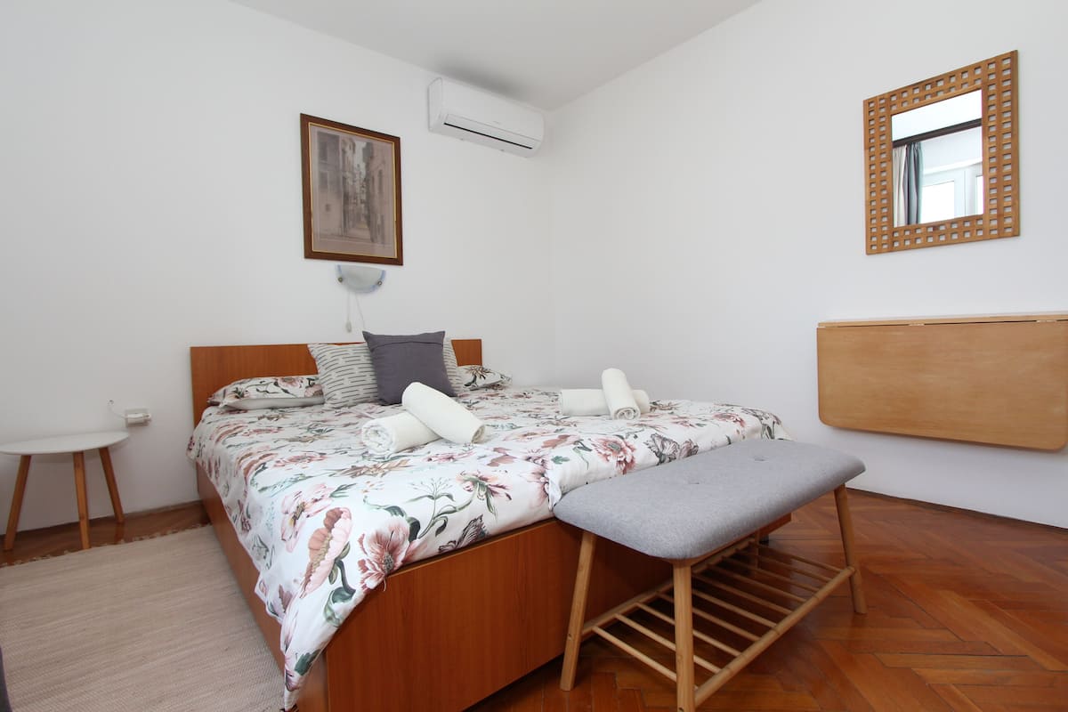 Quarto com cama de madeira, mesa de cabeceira, um pequeno banquinho, ar-condicionado fixado na parede, um quadro emoldurado e um espelho.