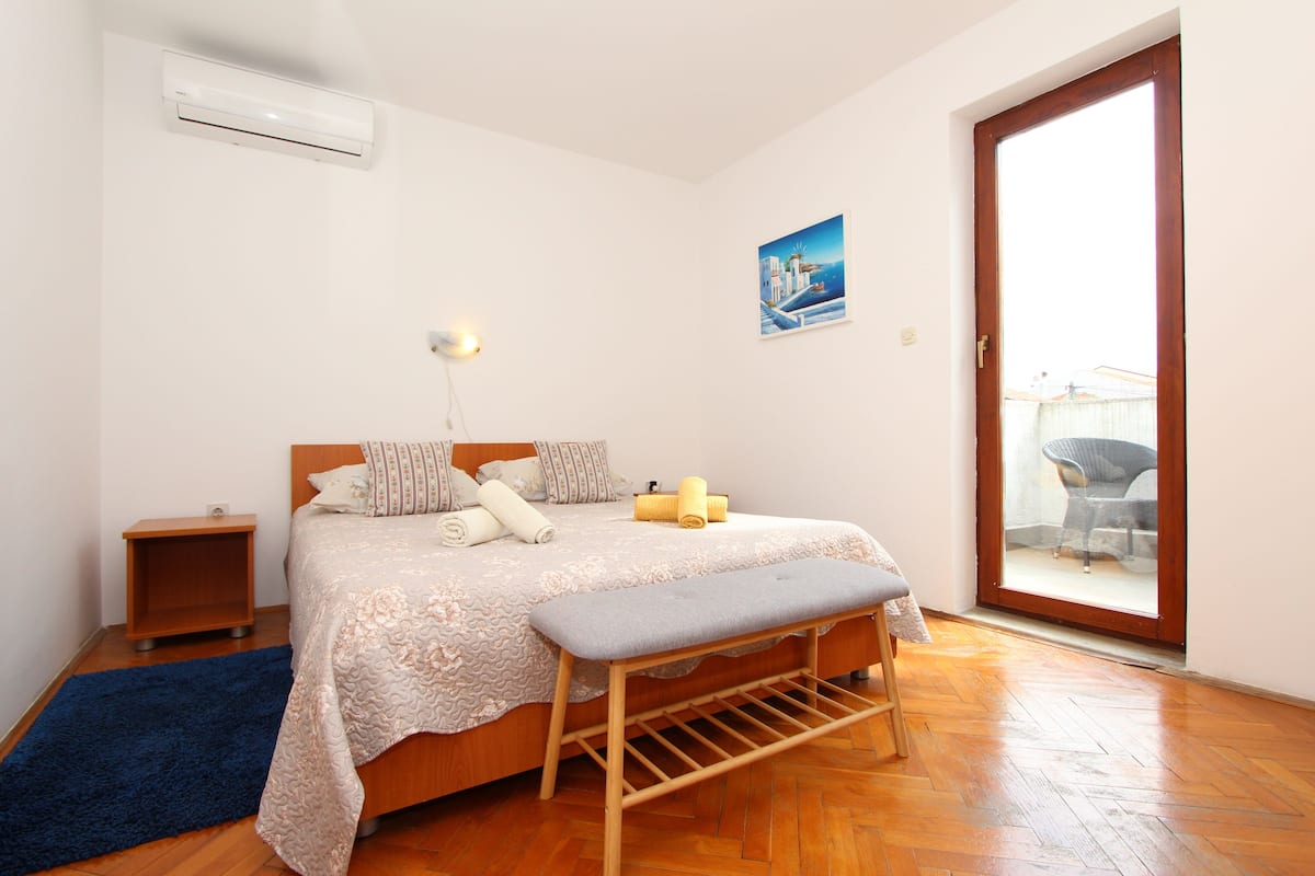 Quarto com cama, mesa de cabeceira, banqueta e vista por uma porta.