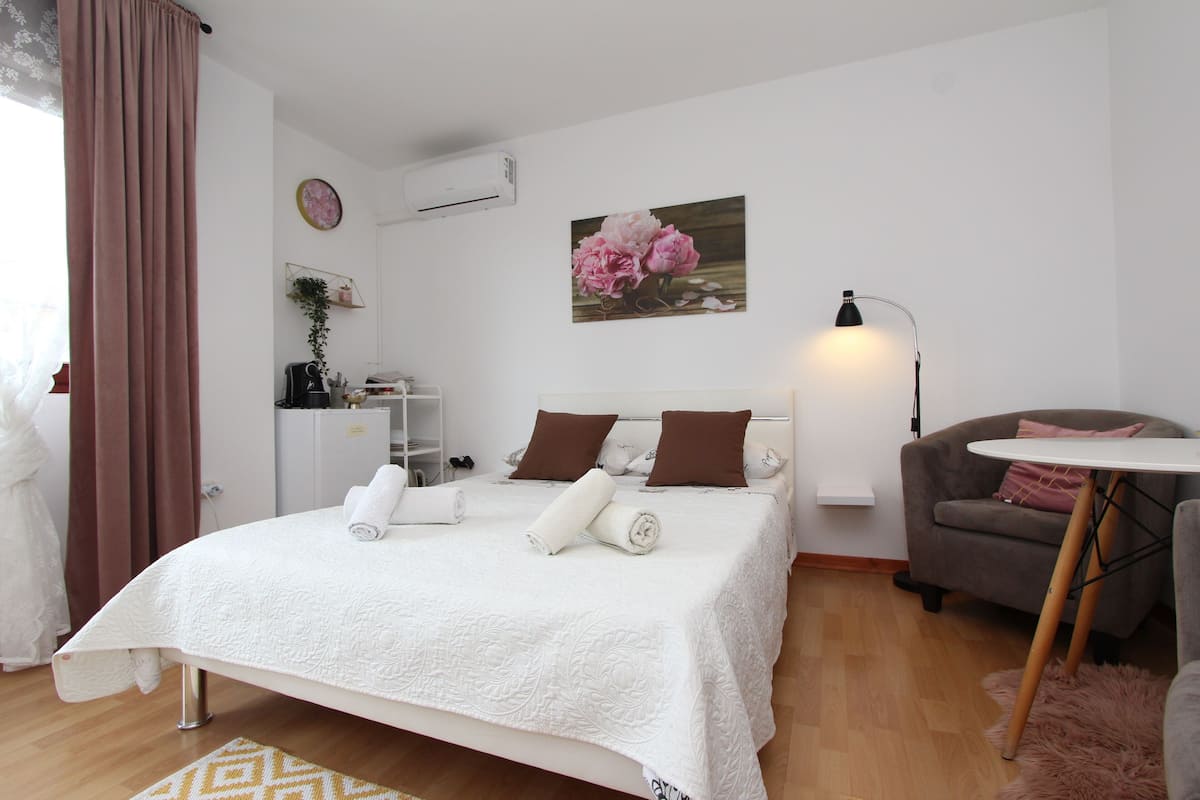 Cama bem arrumada com toalhas enroladas, uma poltrona cinza, uma pequena mesa redonda e um ar condicionado fixado na parede.
