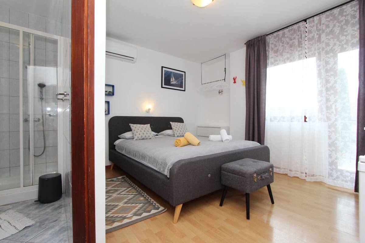 Um quarto moderno com chuveiro, uma cama com cabeceira cinza, mesa de cabeceira e uma janela com cortinas.