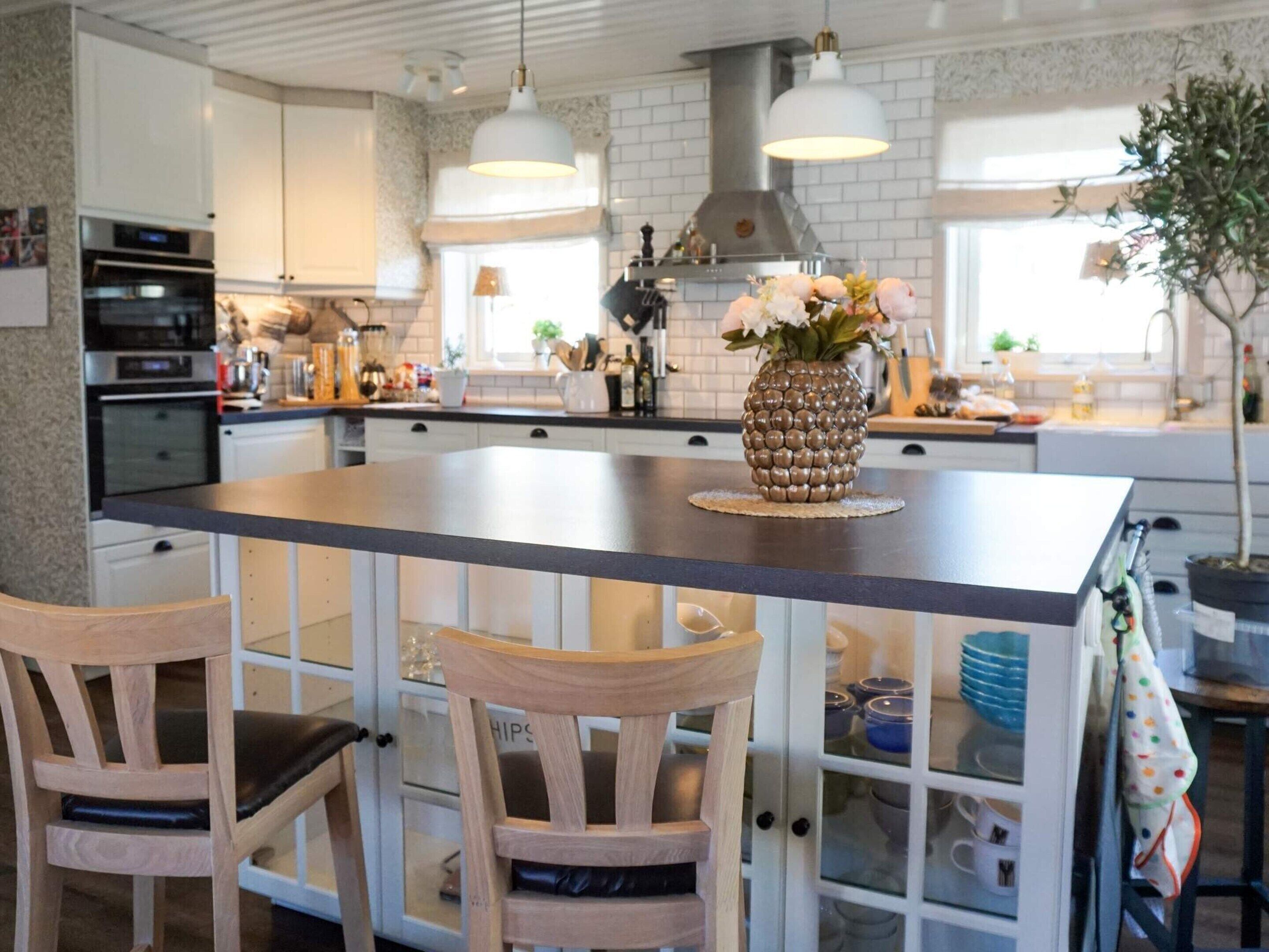 Uma cozinha com armários brancos, uma ilha central com cadeiras, luminárias pendentes e um vaso decorativo de abacaxi.