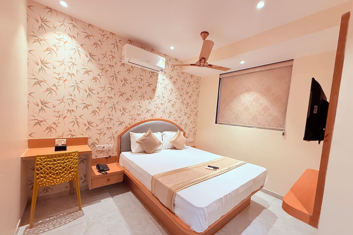 Quarto de hotel com cama, mesa de cabeceira, aparelho de ar condicionado, ventoinha no teto e televisão na parede.