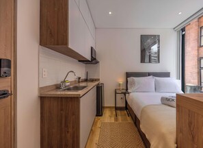 Quarto de hotel compacto com cozinha compacta, cama com roupa de cama branca, mesa de cabeceira pequena com abajur e vista para um edifício de tijolos pela janela.