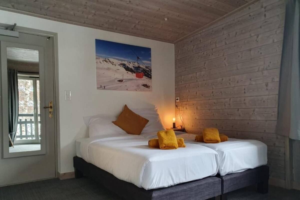 Uma cama de casal com roupa de cama branca e duas almofadas, teto de madeira, pintura de uma pista de esqui e uma porta com vista.