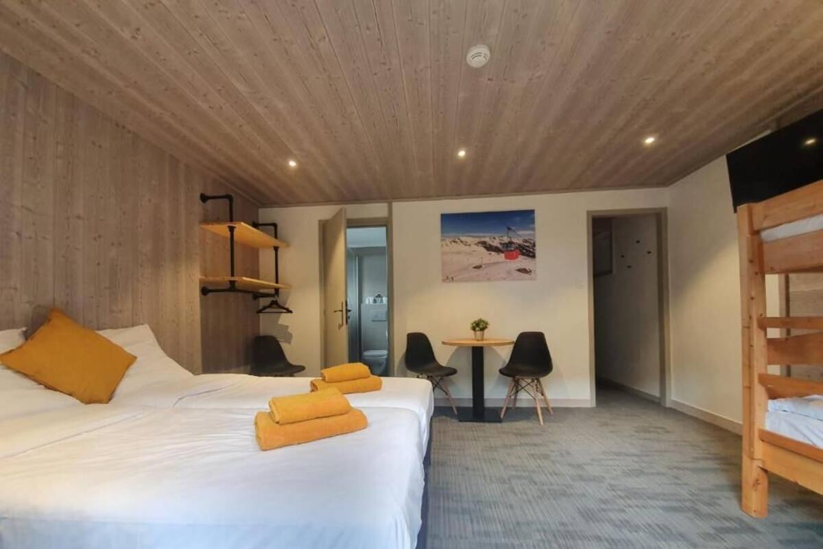 Quarto de hotel com uma cama grande, uma mesa pequena com duas cadeiras, uma cama beliche, paredes de madeira e teto com iluminação embutida.