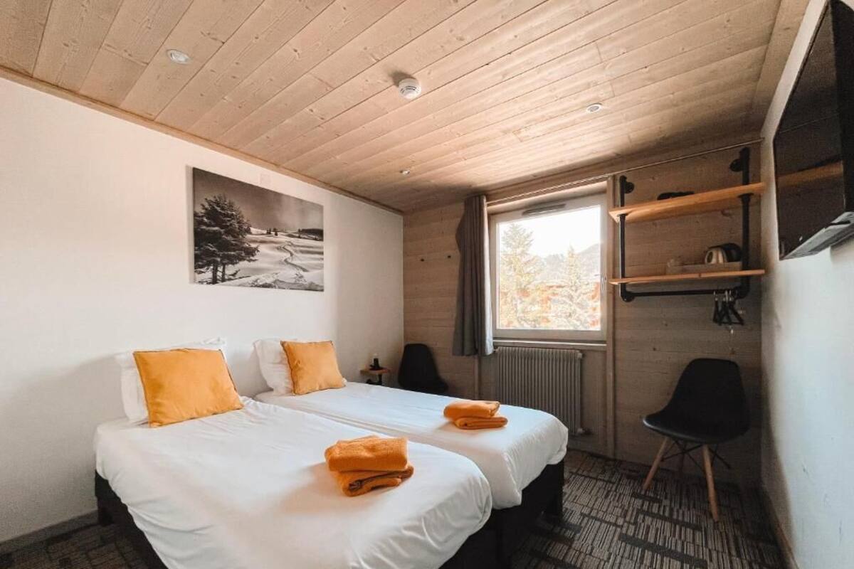 Quarto de hotel com duas camas, uma janela, um radiador, uma cadeira e uma estante com objetos.