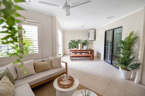 Uma sala de estar com sofá, mesa de centro, ventilador de teto, mesinha de madeira e plantas em vasos.