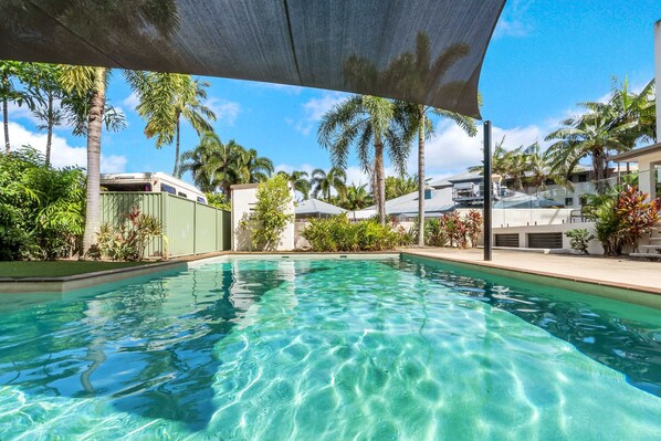 Piscina cristalina com área sombreada, cercada por palmeiras e edifícios modernos.