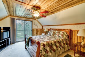 Quarto com teto de madeira, cama com colcha, televisão, abajur e vista para o exterior.