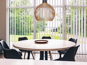 Mesa de jantar redonda com seis cadeiras, luminária pendente e vista para uma paisagem verde através de grandes janelas.