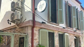 Um edifício com uma placa indicando 'Chori Avau', aparelhos de ar condicionado, persianas verdes e uma rua de paralelepípedos.