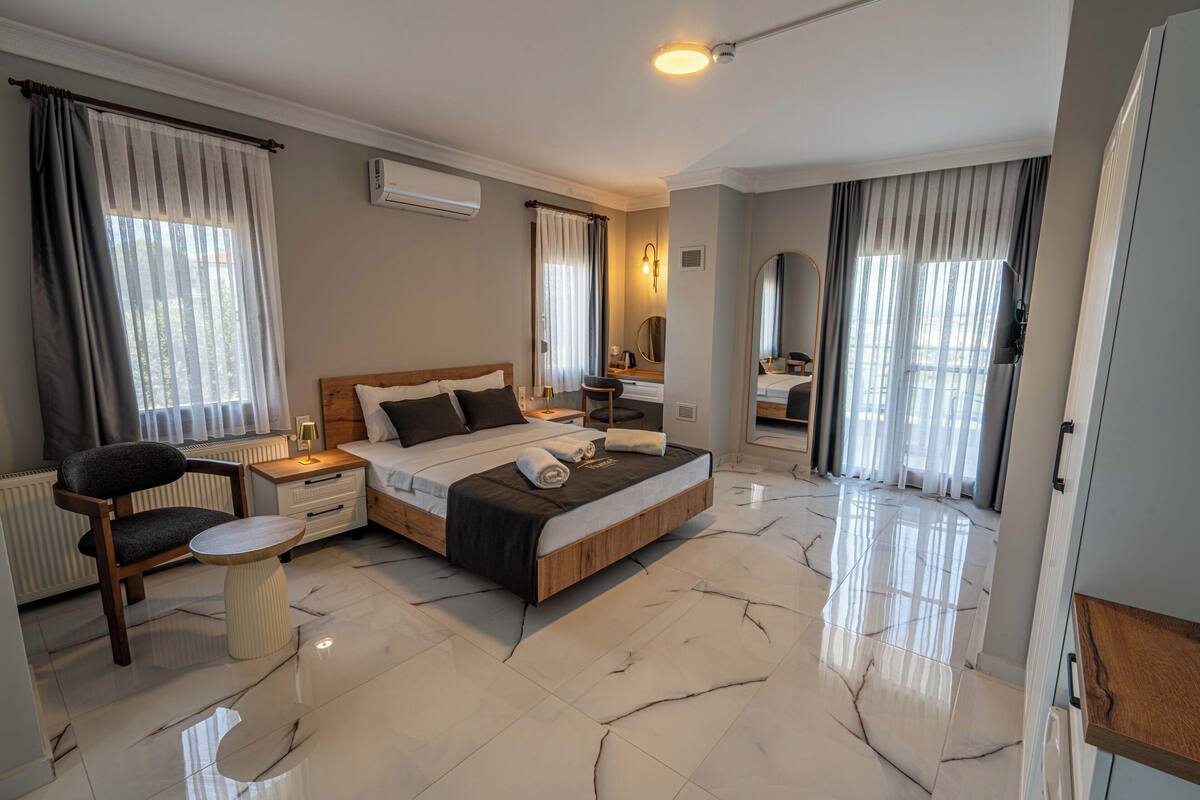 Quarto de hotel moderno com uma cama grande, uma mesinha de cabeceira, uma cadeira, uma pequena mesa redonda e um espelho.