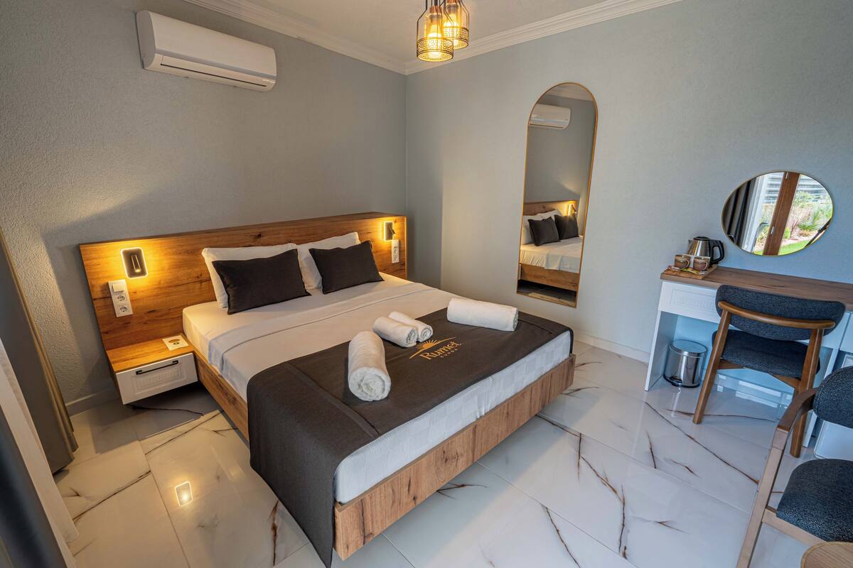 Quarto de hotel com cama, mesa de cabeceira, espelho, escrivaninha, cadeira e um cesto de lixo pequeno.