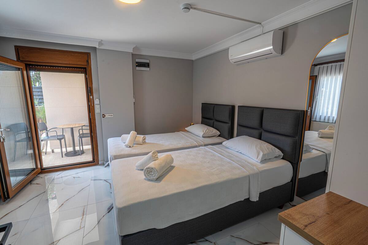 Quarto de hotel com duas camas, uma mesinha, uma cadeira e porta de vidro deslizante para uma área externa.