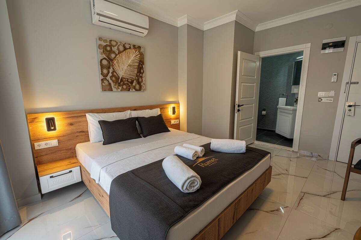 Quarto de hotel moderno com uma cama grande, mesa de cabeceira, ar condicionado fixado na parede e um banheiro visível através de uma porta aberta.