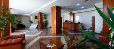 Um saguão de hotel com balcão de recepção, sofás de couro, mesa de centro e escadaria para outro andar.