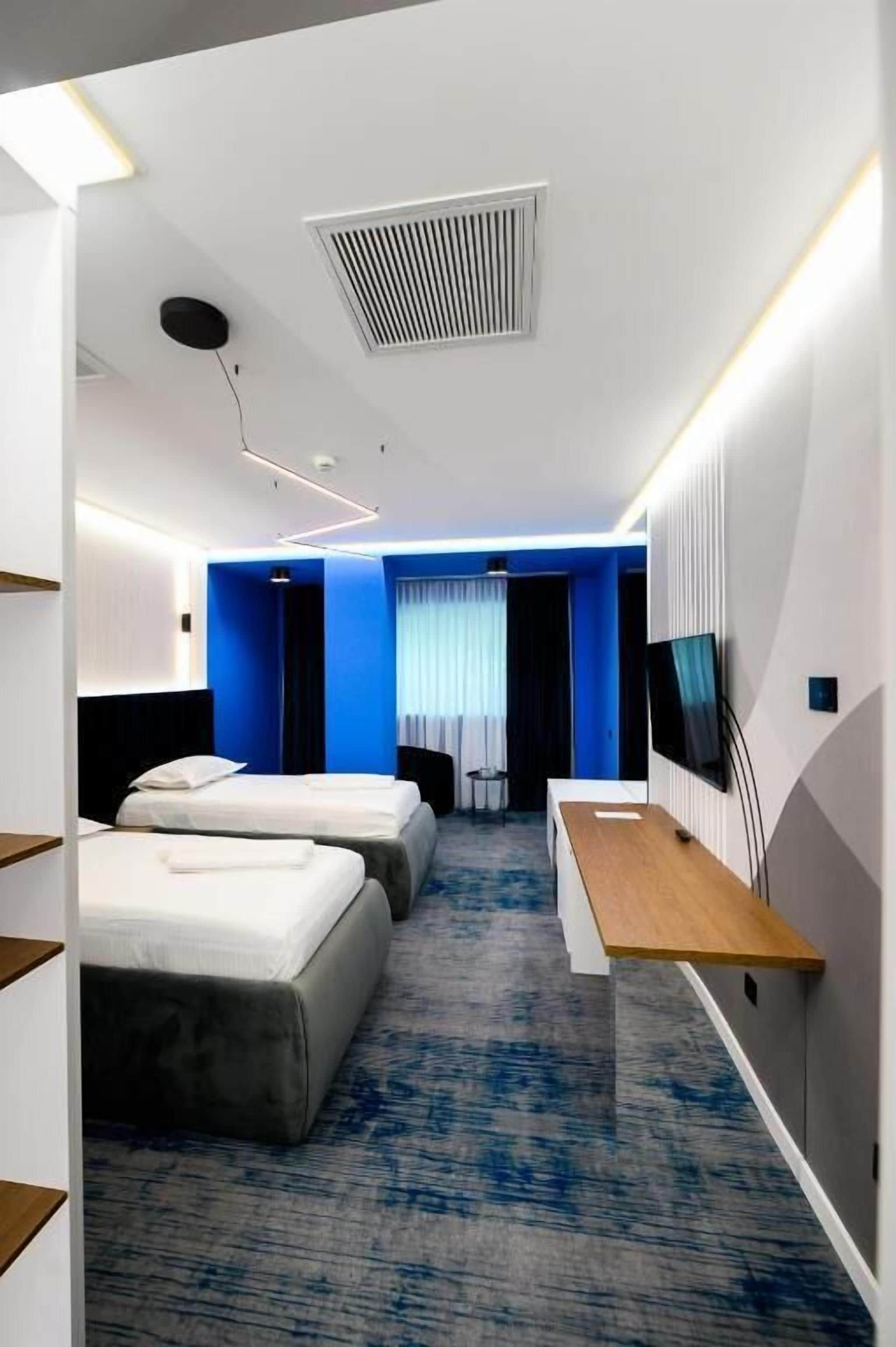 Quarto de hotel com duas camas, uma escrivaninha, uma televisão e um respiradouro no teto.