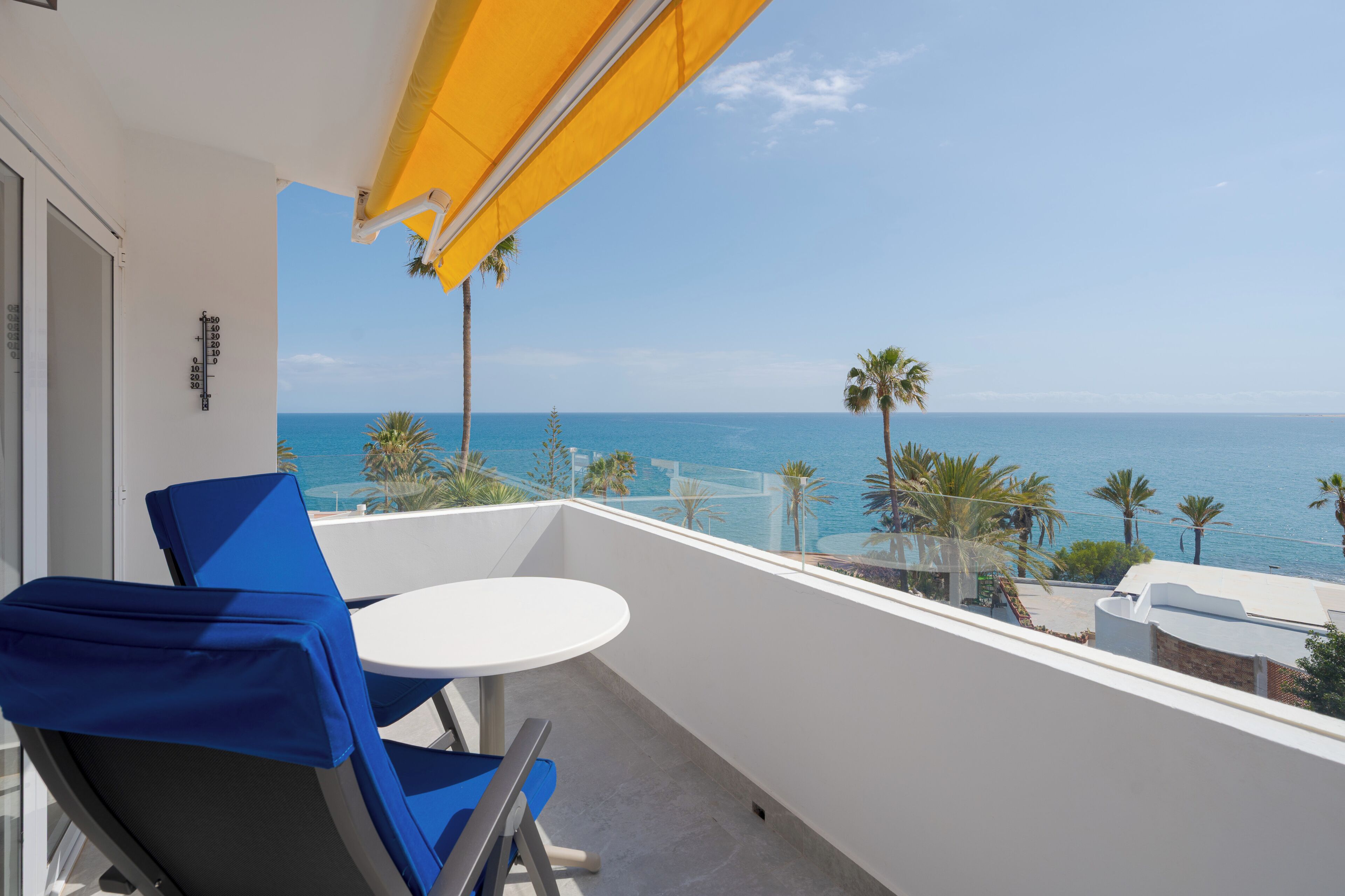 Varanda com mesa branca e cadeiras azuis, com vista para um mar azul cristalino e palmeiras.