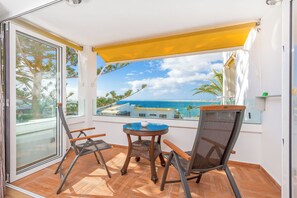 Varanda com mesa e cadeiras, toldo amarelo e vista para o mar e edifícios.