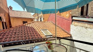 Uma varanda com mesa de vidro, duas cadeiras, um guarda-sol listrado em azul e um telhado de azulejos ao fundo.