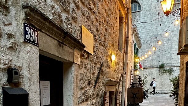 Uma rua de pedra estreita com a entrada de um edifício marcada '396', uma porta de madeira e uma pequena área externa com mesas e cadeiras.