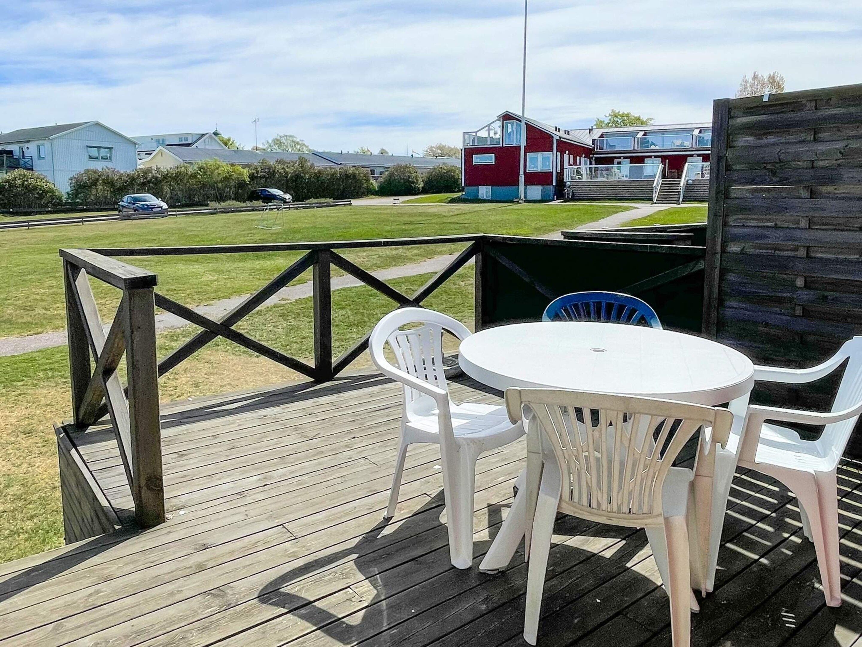 Um deck de madeira com uma mesa e cadeiras brancas, com vista para uma área gramada e edifícios ao fundo.