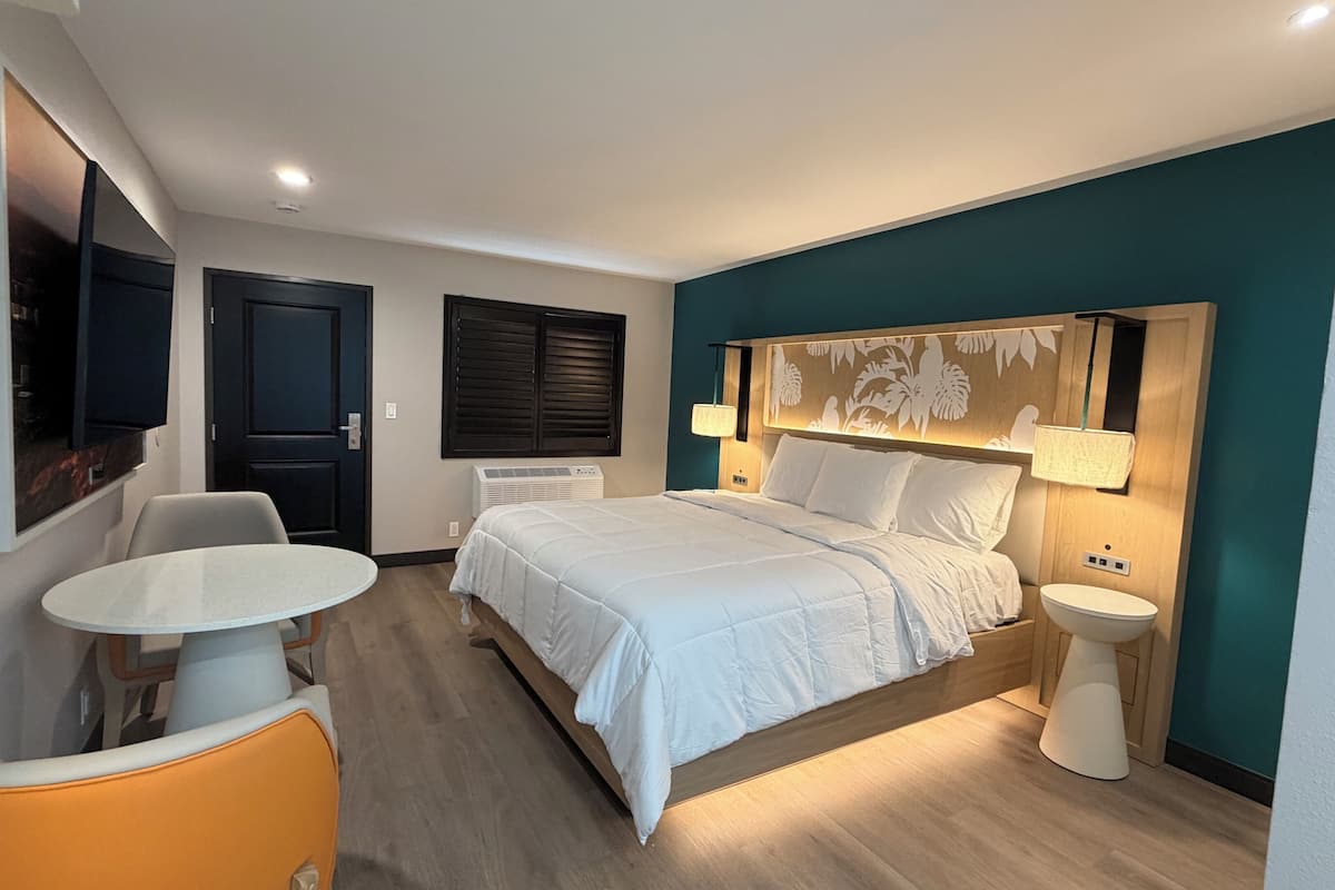 Quarto de hotel com cama, escrivaninha, cadeira, televisão e janela com persianas.
