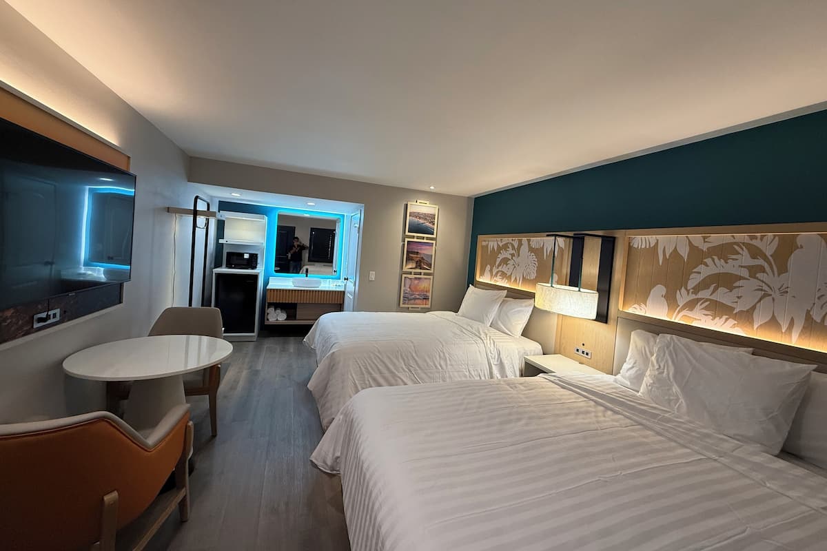 Quarto de hotel com duas camas, televisão, escrivaninha e banheiro com espelho.