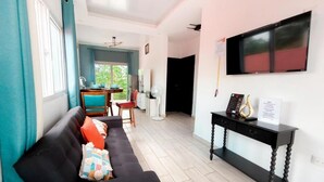 Uma sala de estar com um sofá preto, uma mesa de jantar com cadeiras, uma TV na parede e uma pequena mesa com um ventilador.