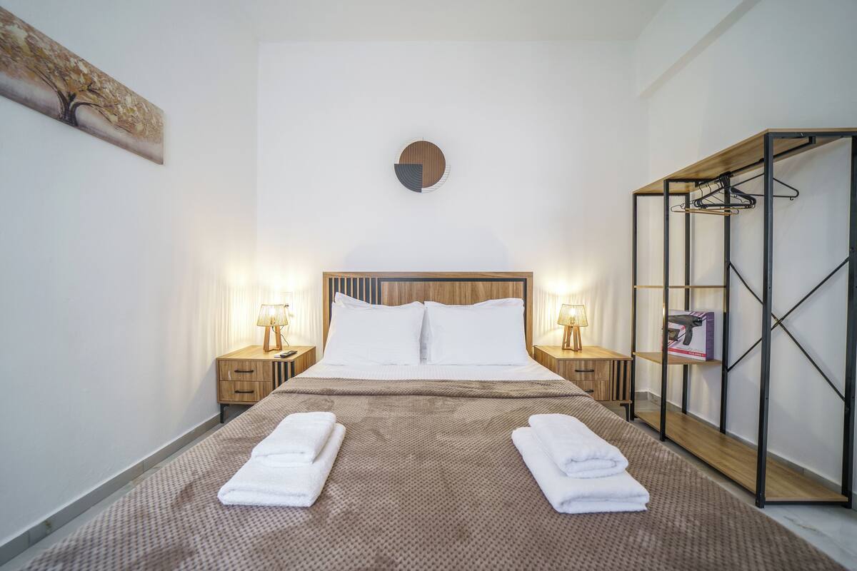 Quarto de hotel com cama, duas mesas de cabeceira com abajures, um guarda-roupa e prateleira na parede.