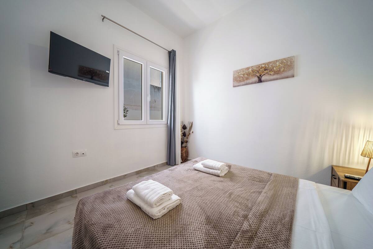 Quarto com cama, televisão fixada na parede, janela com cortinas e um quadro na parede.
