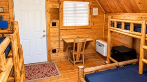 Uma cabana com interior em madeira, uma porta, uma janela com persianas, uma mesa, uma cadeira, uma cama e um aquecedor.