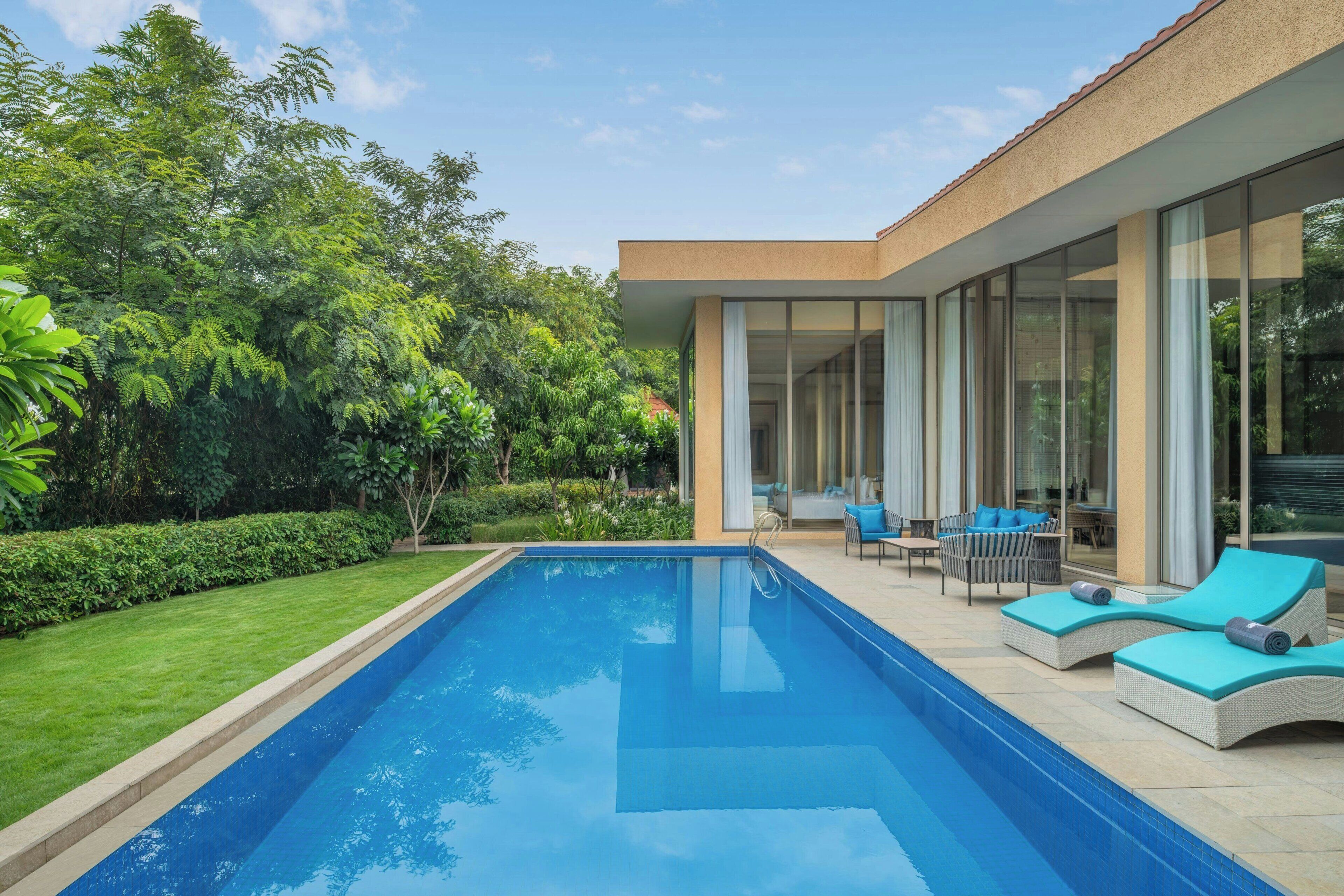 Uma área moderna de piscina ao ar livre, com uma piscina azul translúcida, rodeada por vegetação exuberante e um edifício contemporâneo com grandes janelas.