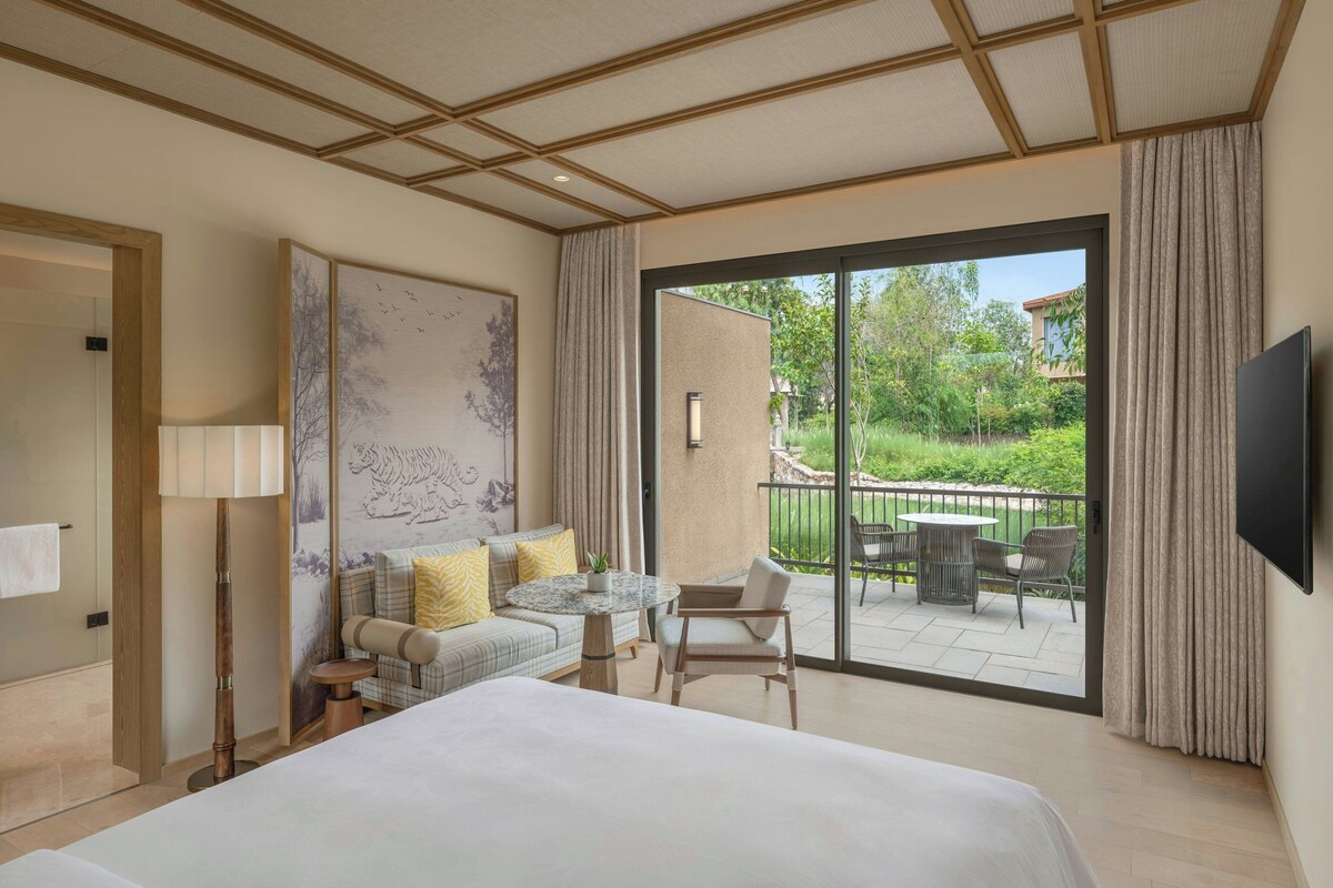 Quarto de hotel com uma cama grande, um sofá com almofadas, uma mesa pequena, uma cadeira, uma TV e uma varanda com mobiliário de exterior.