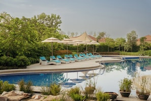 Área de piscina com espreguiçadeiras e guarda-sóis, rodeada por vegetação e um edifício ao fundo.