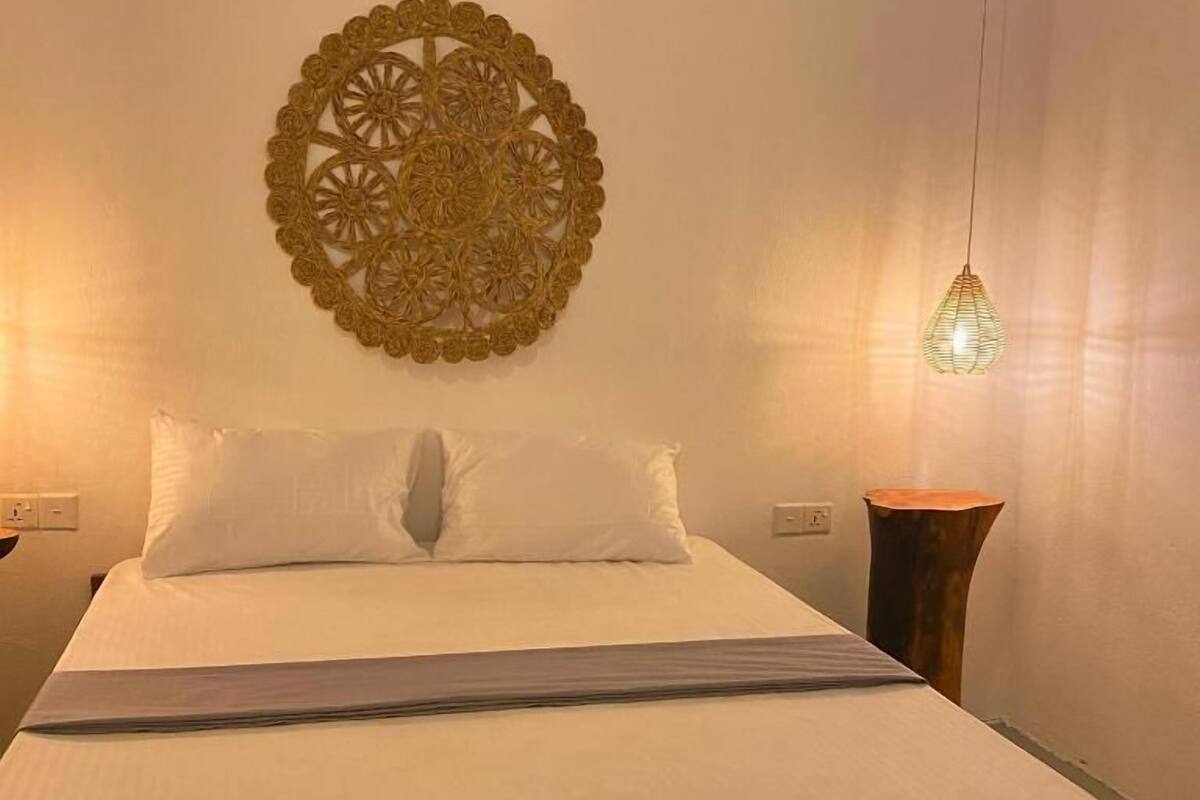 Cama bem arrumada com colchão e almofadas brancas, um painel decorativo acima e uma luminária pendente à direita.