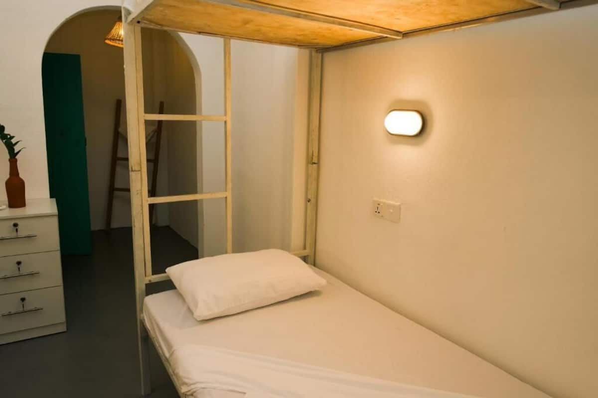 Uma cama de solteiro com travesseiro branco, luminária na parede e cômoda branca com gavetas.