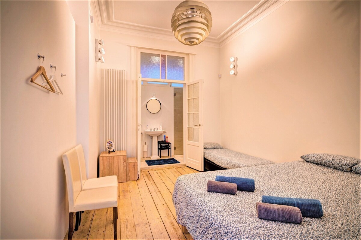 Quarto com cama, cadeira, uma mesinha, radiador e porta que dá para outro espaço.
