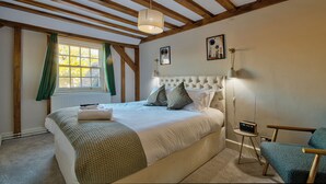 Quarto com cama grande, mesa de cabeceira, cadeira, janela com cortinas e teto com vigas de madeira.