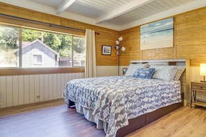 Quarto com cama, mesa de cabeceira, janela com cortinas, ventoinha na parede e pintura com tema praiano.