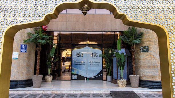 Entrada de hotel com um arco decorativo, uma placa com o nome do hotel e plantas em vasos.