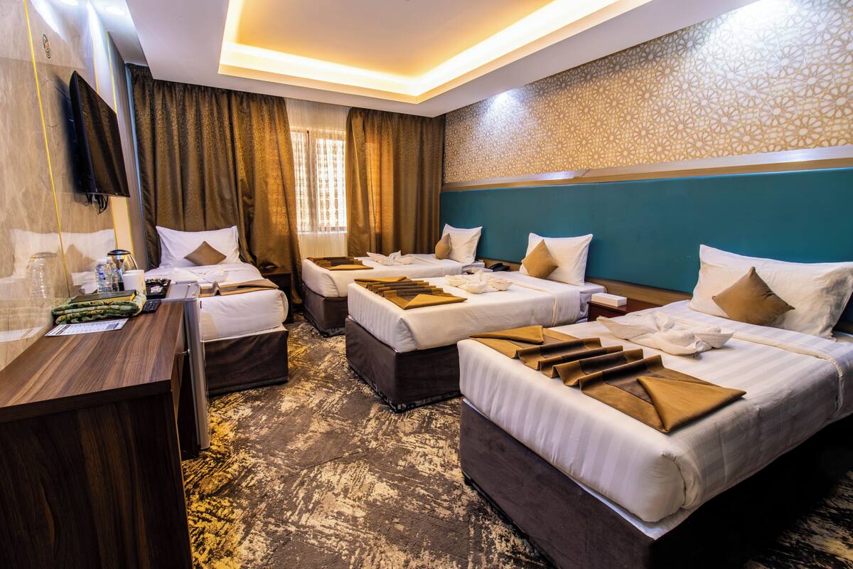 Quarto de hotel com quatro camas alinhadas, uma escrivaninha com telefone e cafeteira, e uma televisão fixada na parede.