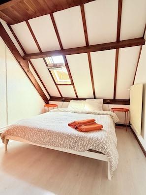 Quarto com teto inclinado, cama com colcha estampada, duas almofadas, mesa de cabeceira e radiador.